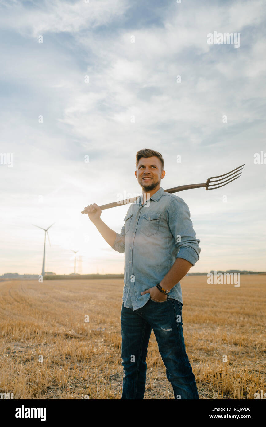 Portrait smiling young man holding pitchfork standing stubble field hi ...