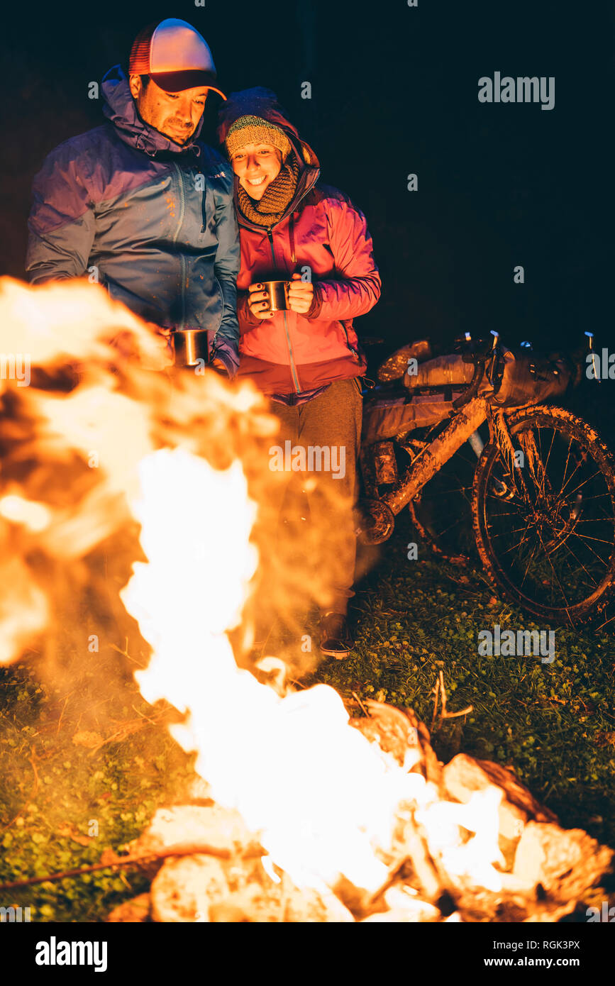 Couple with with bikes standing at camp fire looking at flame by night ...