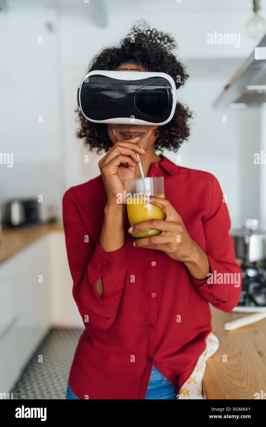 Woman standing in her kitchen, wearing VR goggles, drinking orange ...