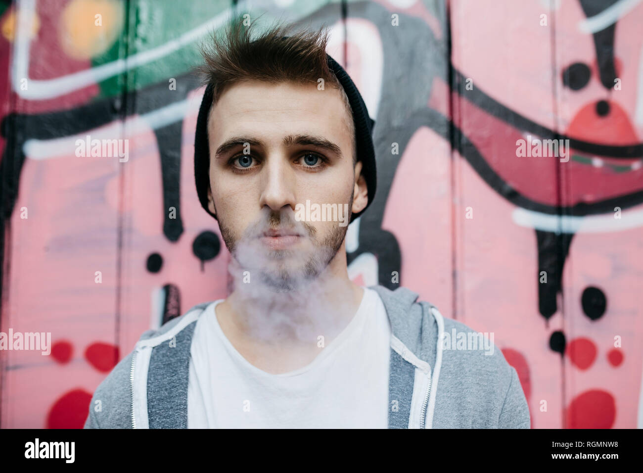 Young man standing in front of graffiti, smoking electronic cigarette ...