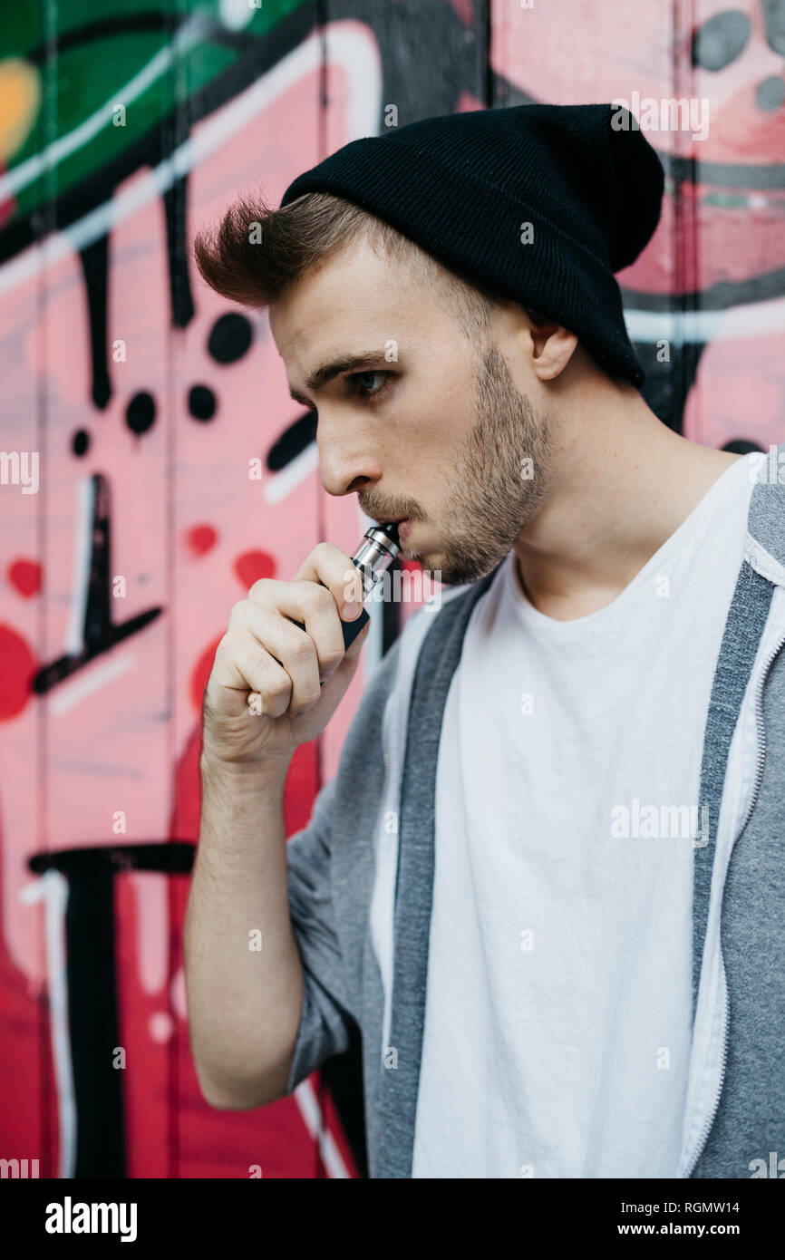 Young man standing in front of graffiti, smoking electronic cigarette ...