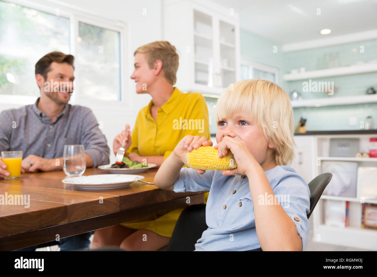 Boy eating corn cob in kitchen with parents in background Stock Photo ...