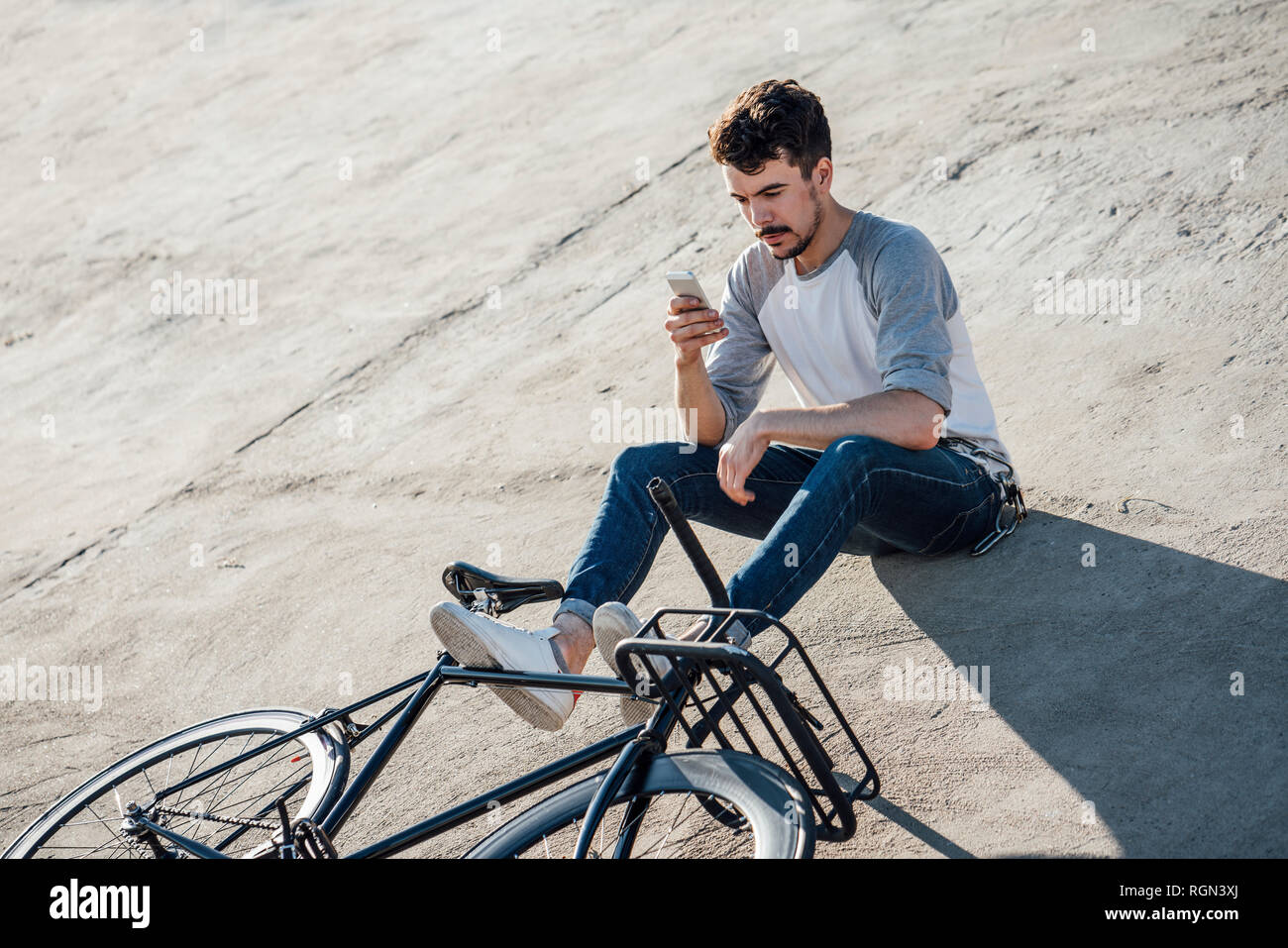 Young man with commuter fixie bike sitting on concrete wall using cell ...