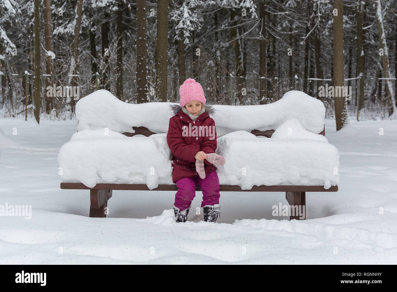A little girl on old wooden bench full of snow near the forest puts ...
