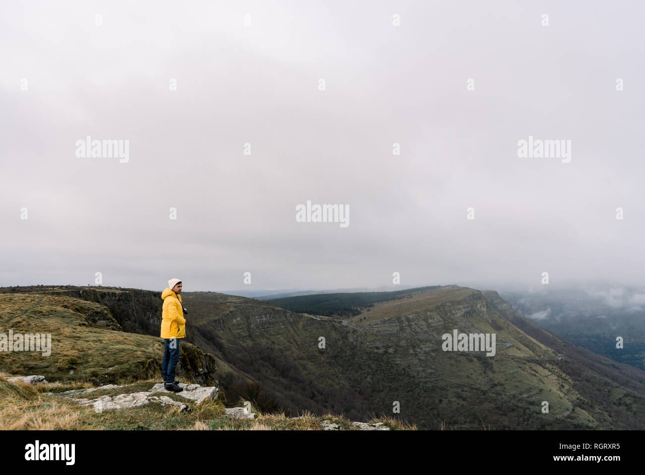 Side view of guy in yellow coat and hat standing on top of mountain and ...