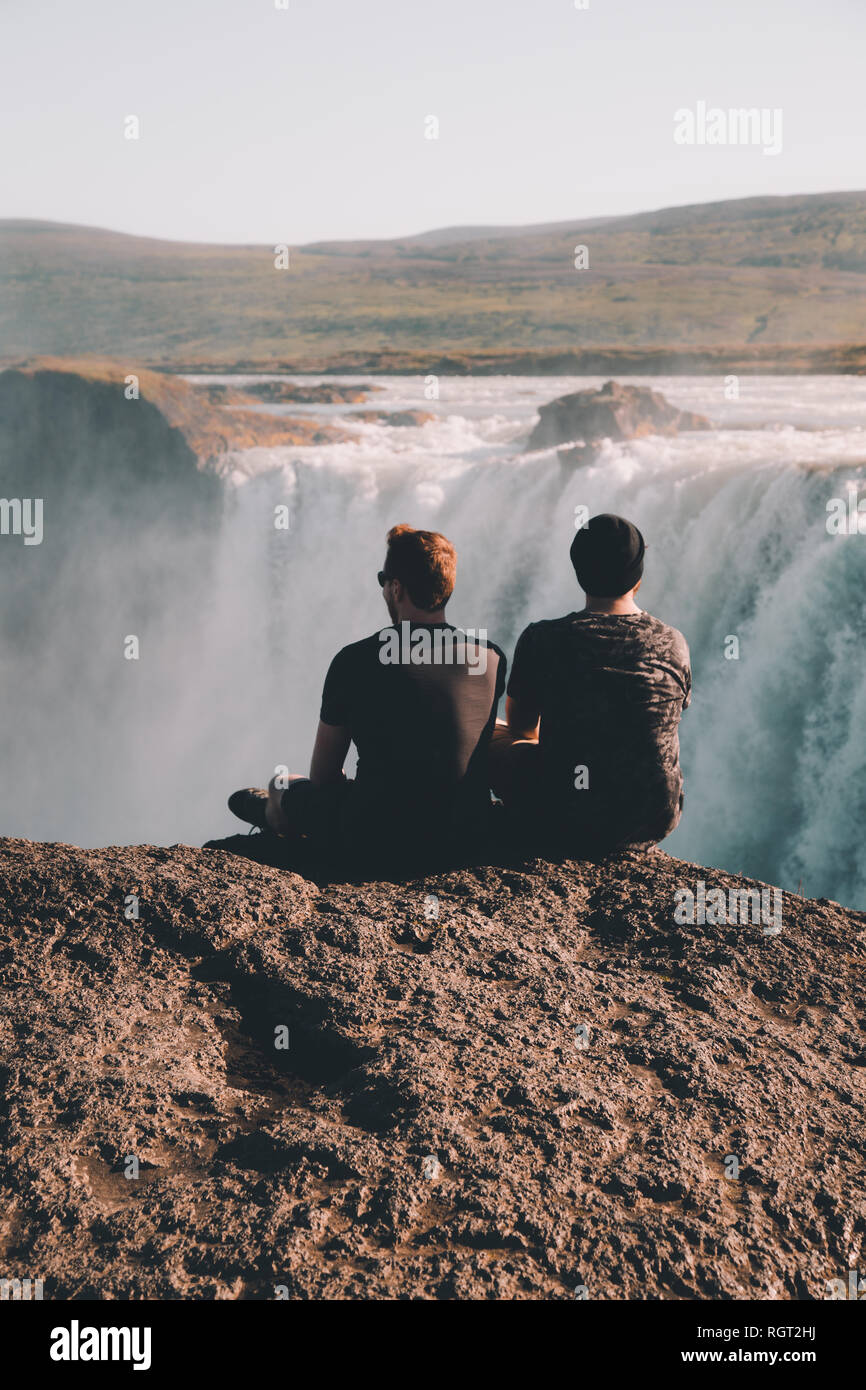 Back view of two men sitting on cliff and admiring amazing waterfall in ...