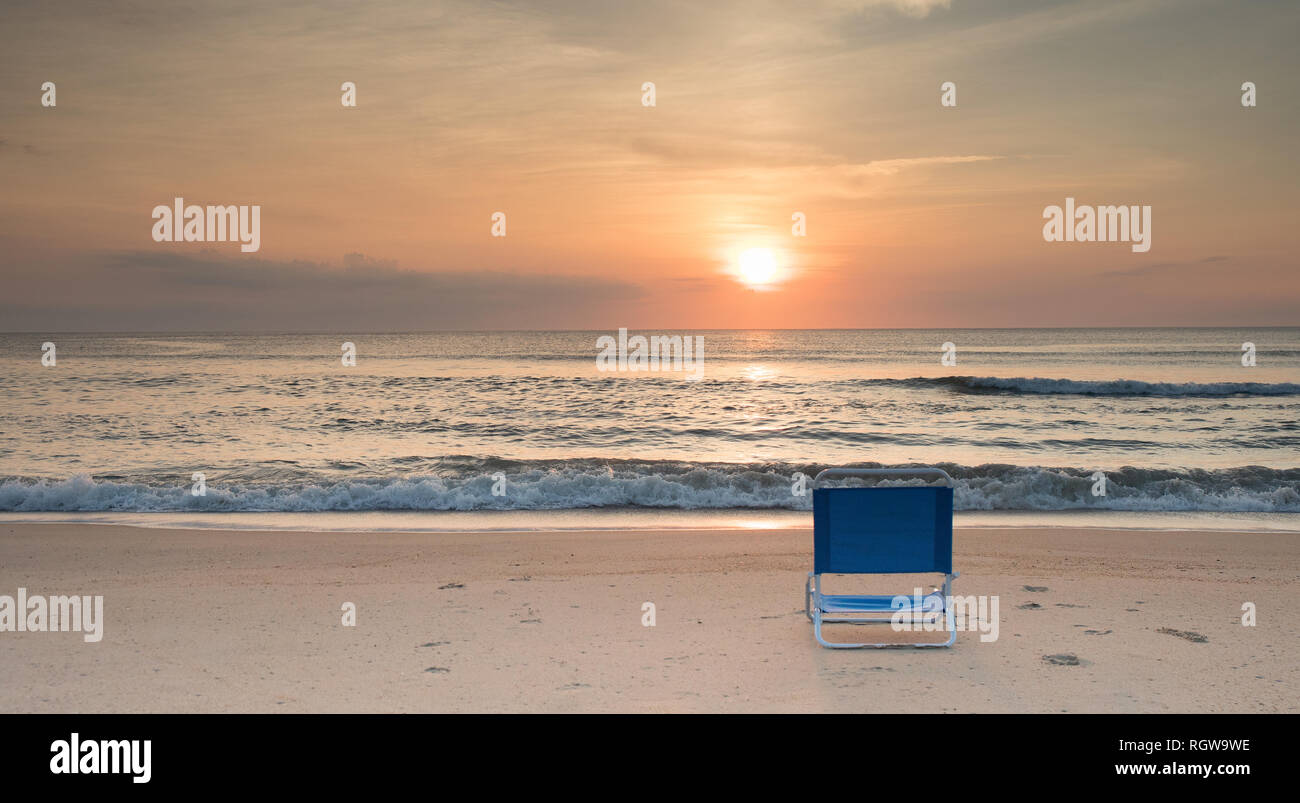 An empty blue beach chair facing the sunrise with waves crashing ashore ...