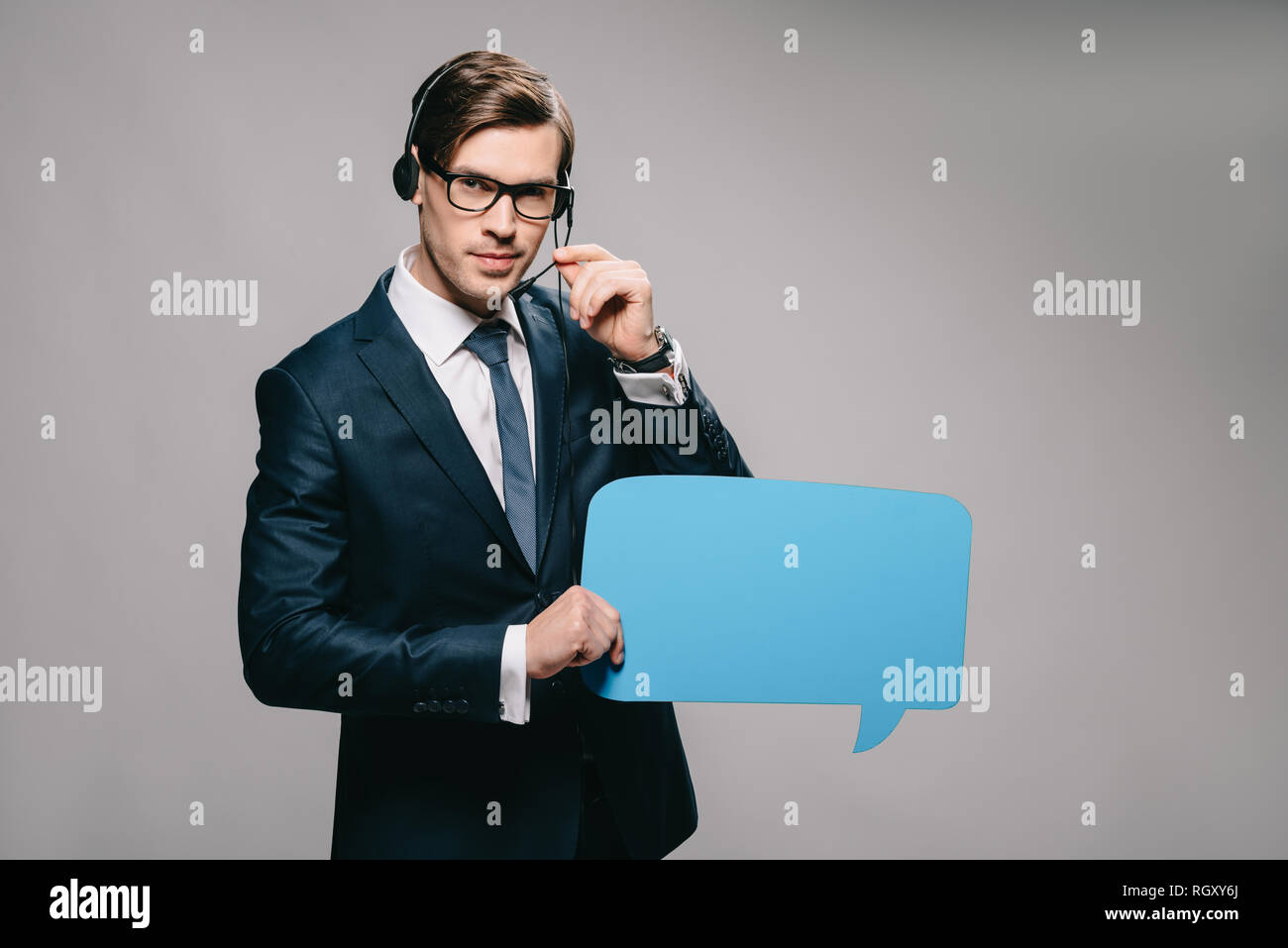 serious man in suit wearing headset and holding speech bubble on grey ...