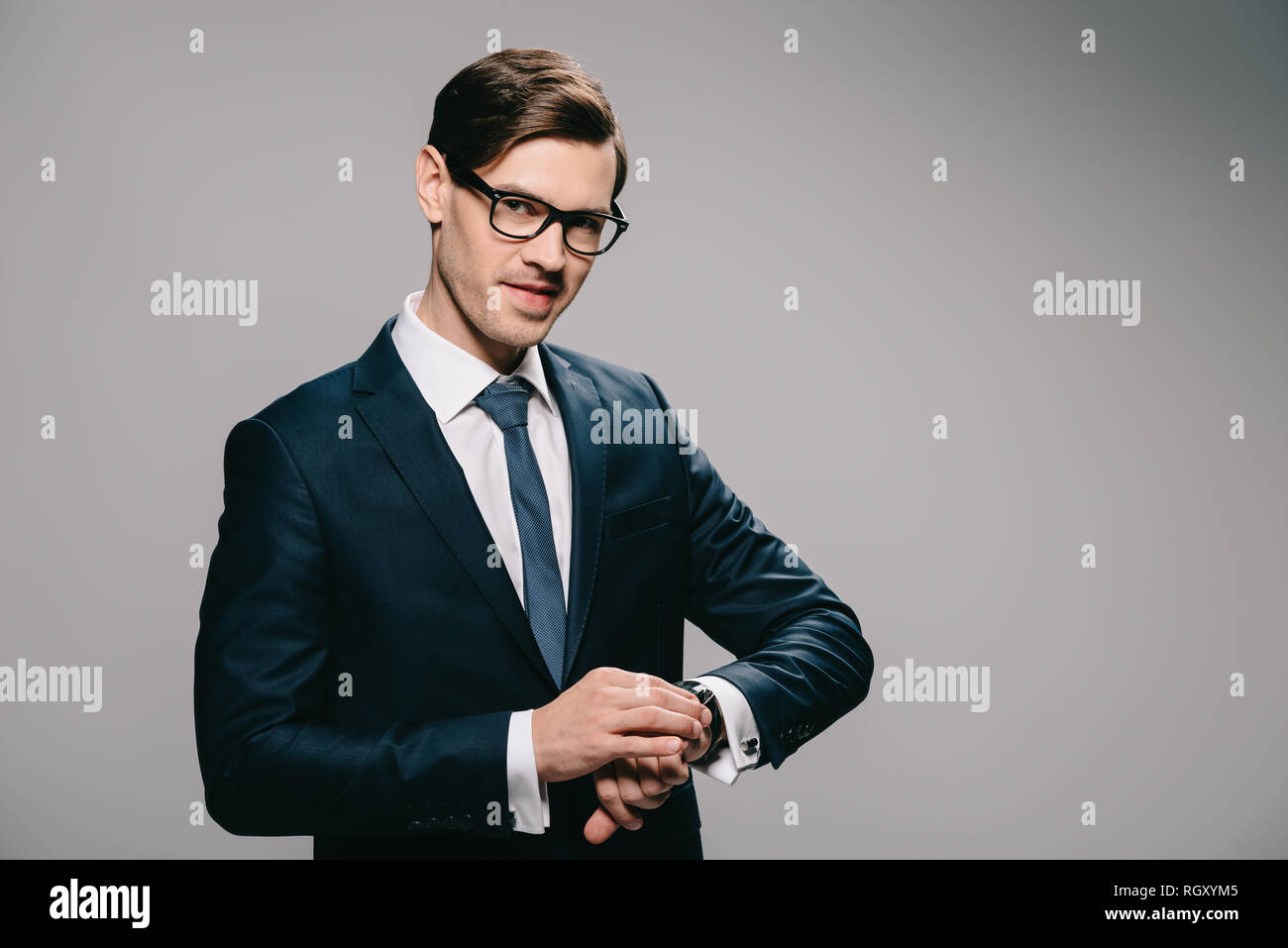 confident man in glasses and suit holding watch on grey background ...