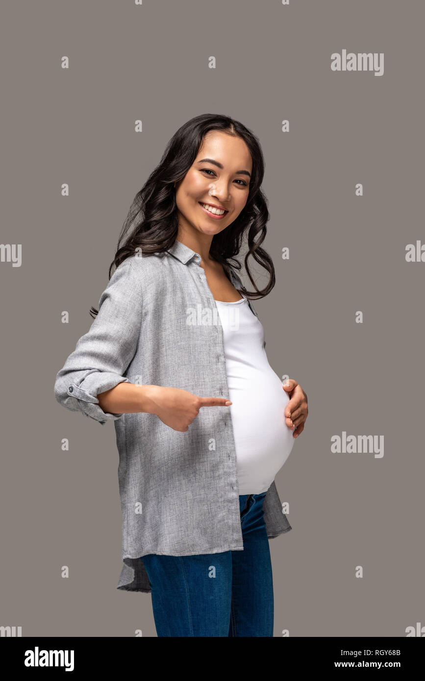 Young pregnant asian woman pointing at tummy and smiling isolated on ...