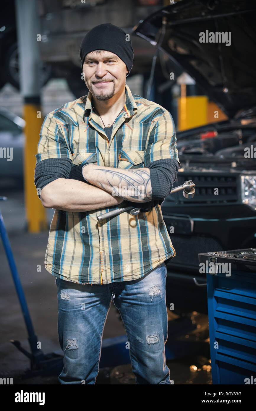Car repair service. Brutal mechanic man stands holding tools and smiles ...
