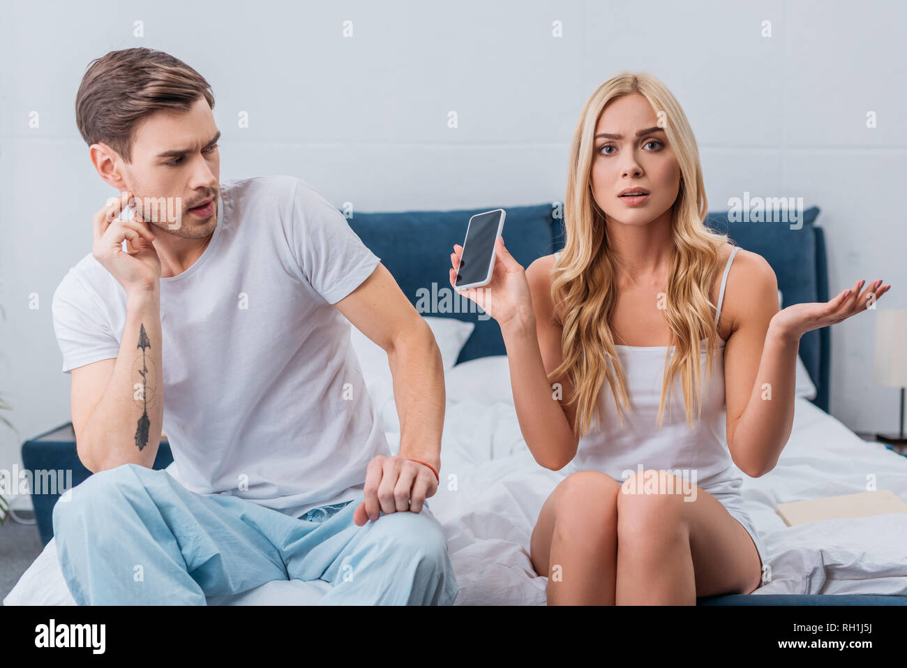 confused woman holding smartphone and looking at camera while sitting ...
