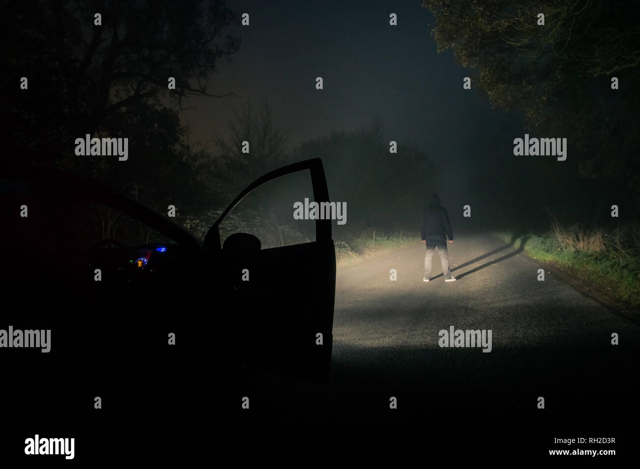 A lone hooded figure standing on an empty misty spooky road at night ...