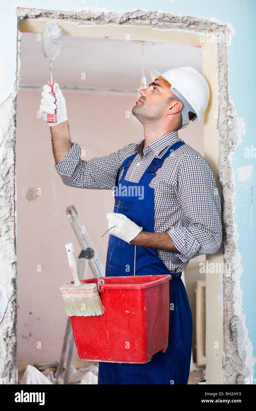 Constructor in helmet repairing wall with roller and bucket indoors ...