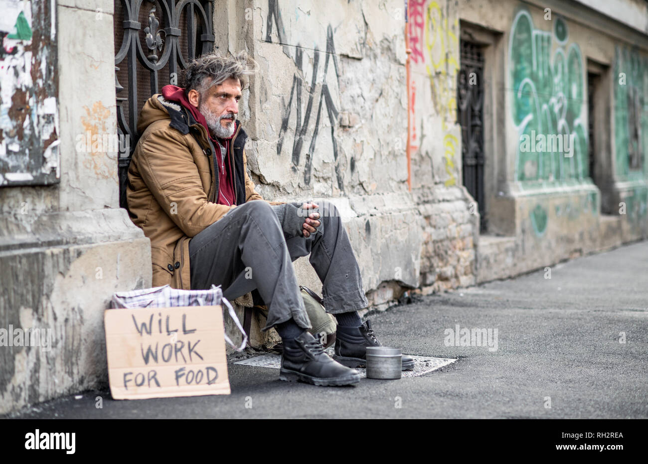 A side view of homeless beggar man sitting in front of gate outdoors in ...