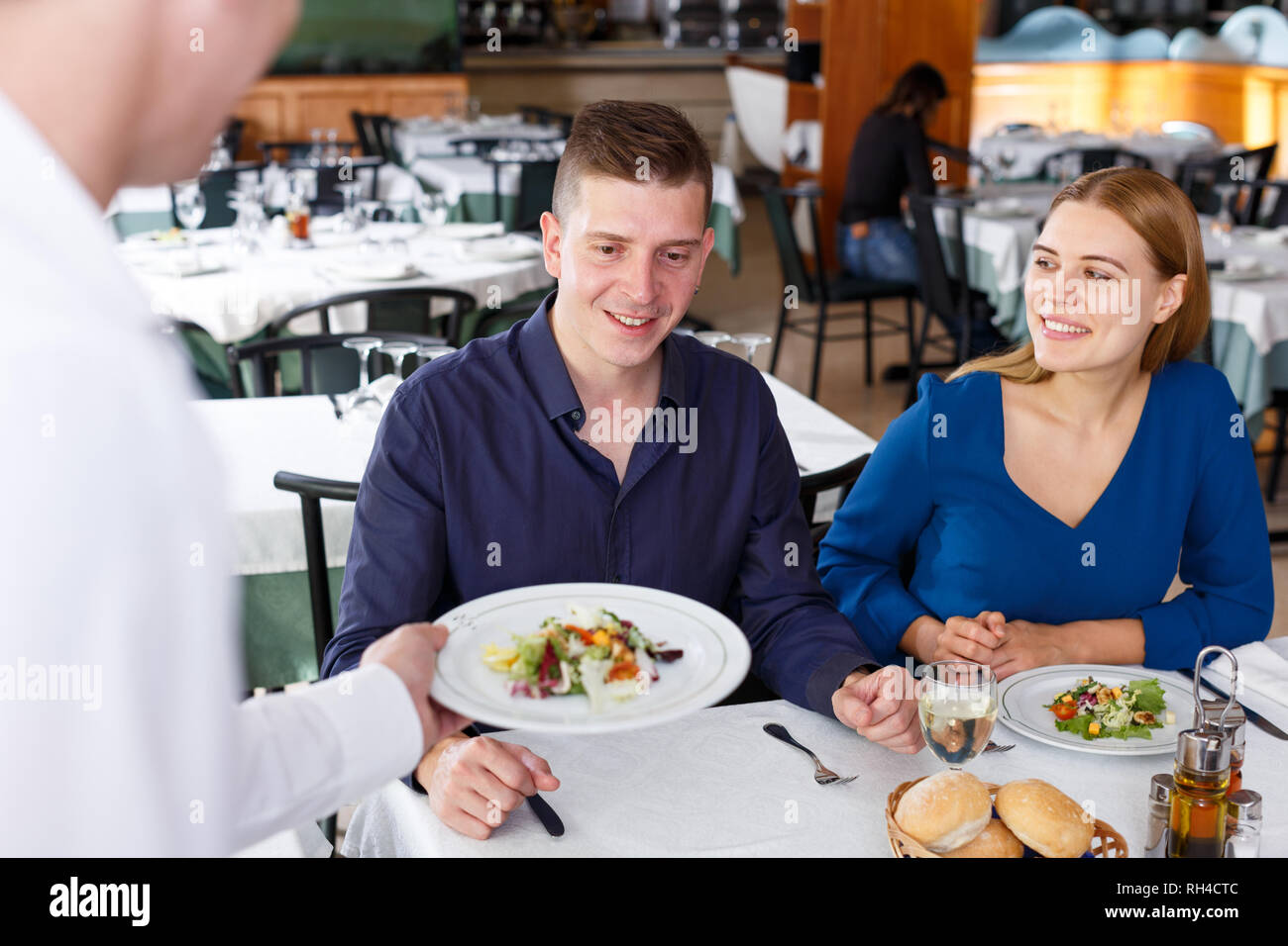 Cheerful waiter serving dishes to adult couple having dinner at ...