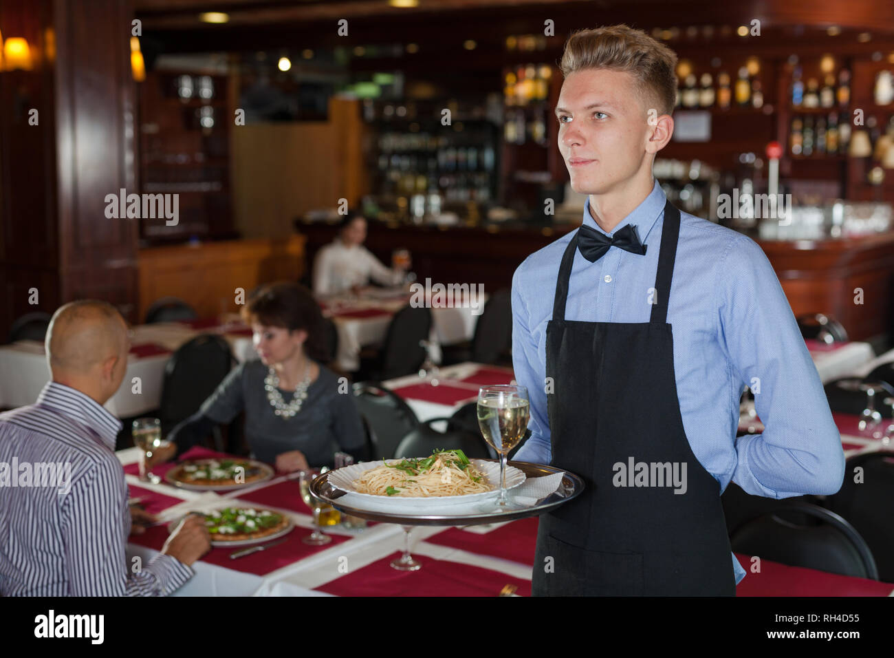 Polite waiter holding tray in restaurant with customers behind him ...