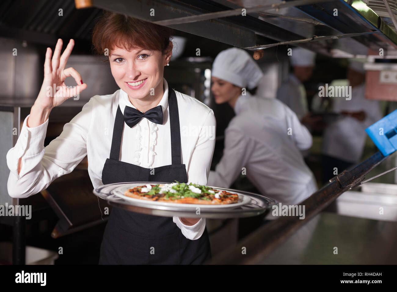 Smiling waitress standing in restaurant kitchen with ordered pizza ...