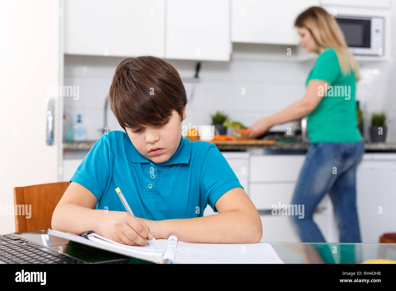 Positive schoolboy doing homework using laptop at kitchen interior ...
