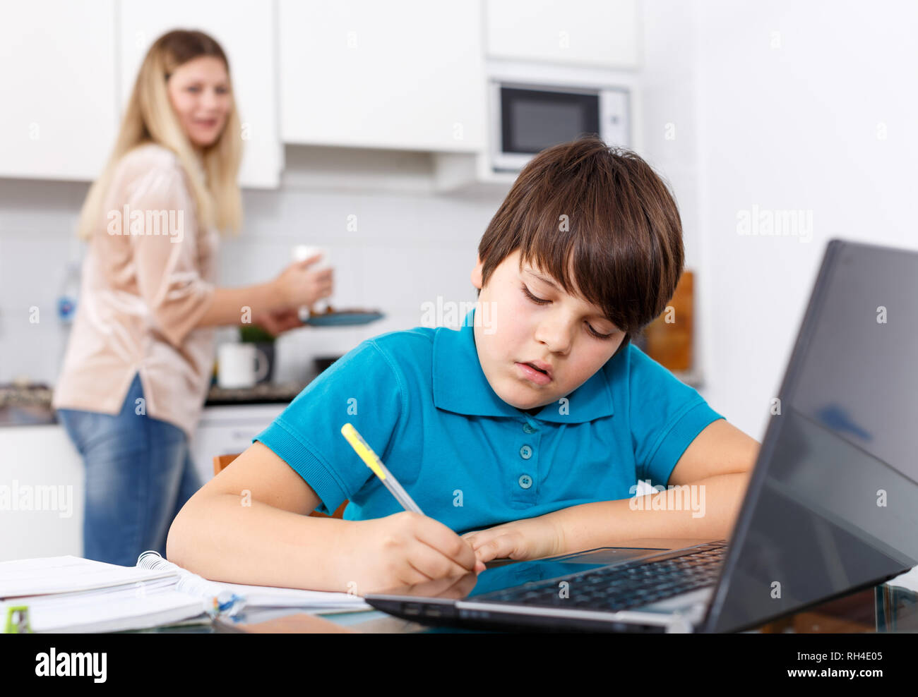 Positive schoolboy doing homework using laptop at kitchen interior ...