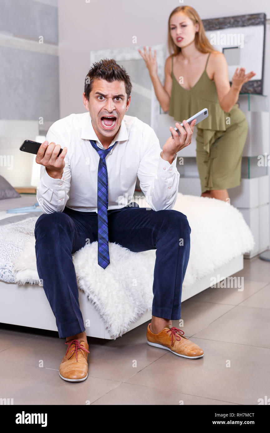 Portrait of angry shouting man sitting with two cell phones on bed in ...