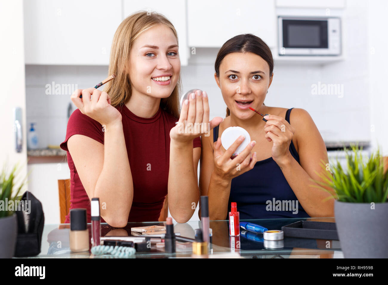Two modern girls applying make up and helping each other at kitchen ...