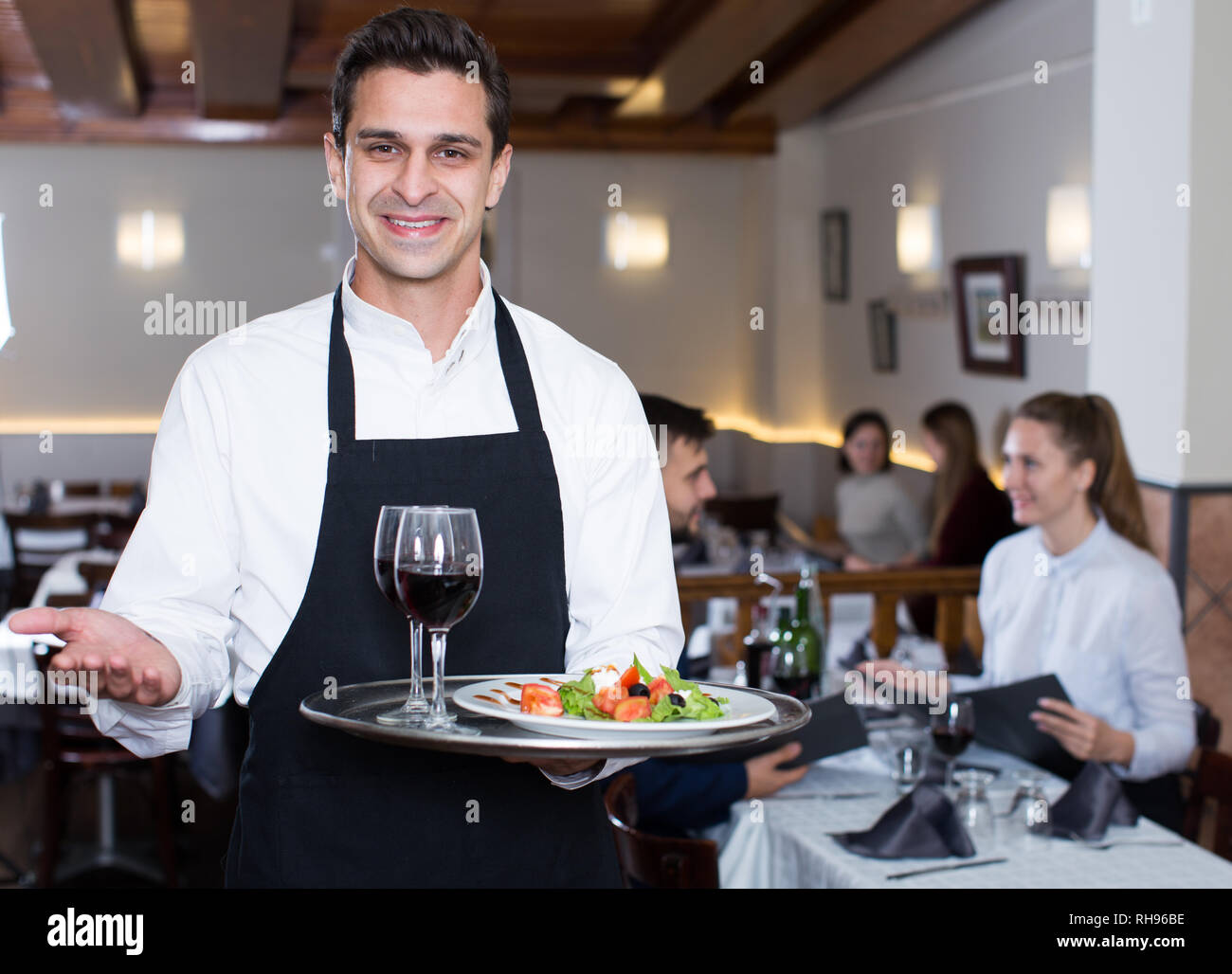 Polite waiter holding tray at restaurant with customers his behind ...