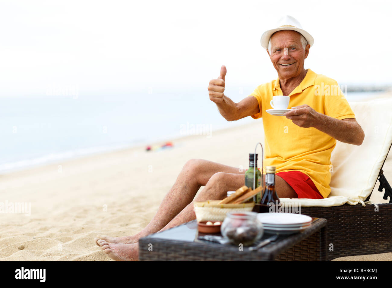 Positive mature man sitting with cup of hot drink on the beach and ...