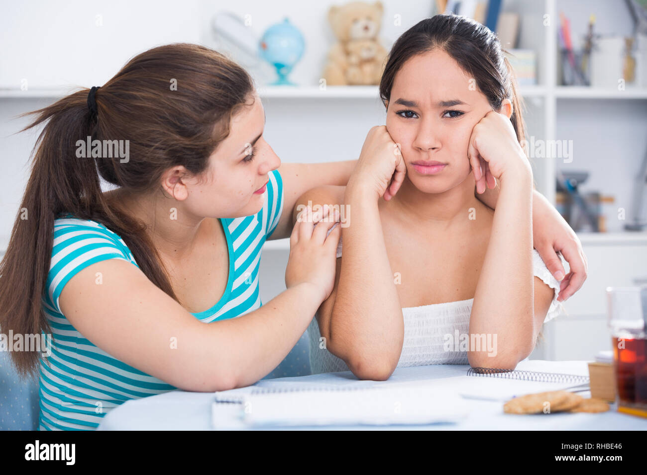 Sad teenager girl sitting at table at home while female friend soothing ...