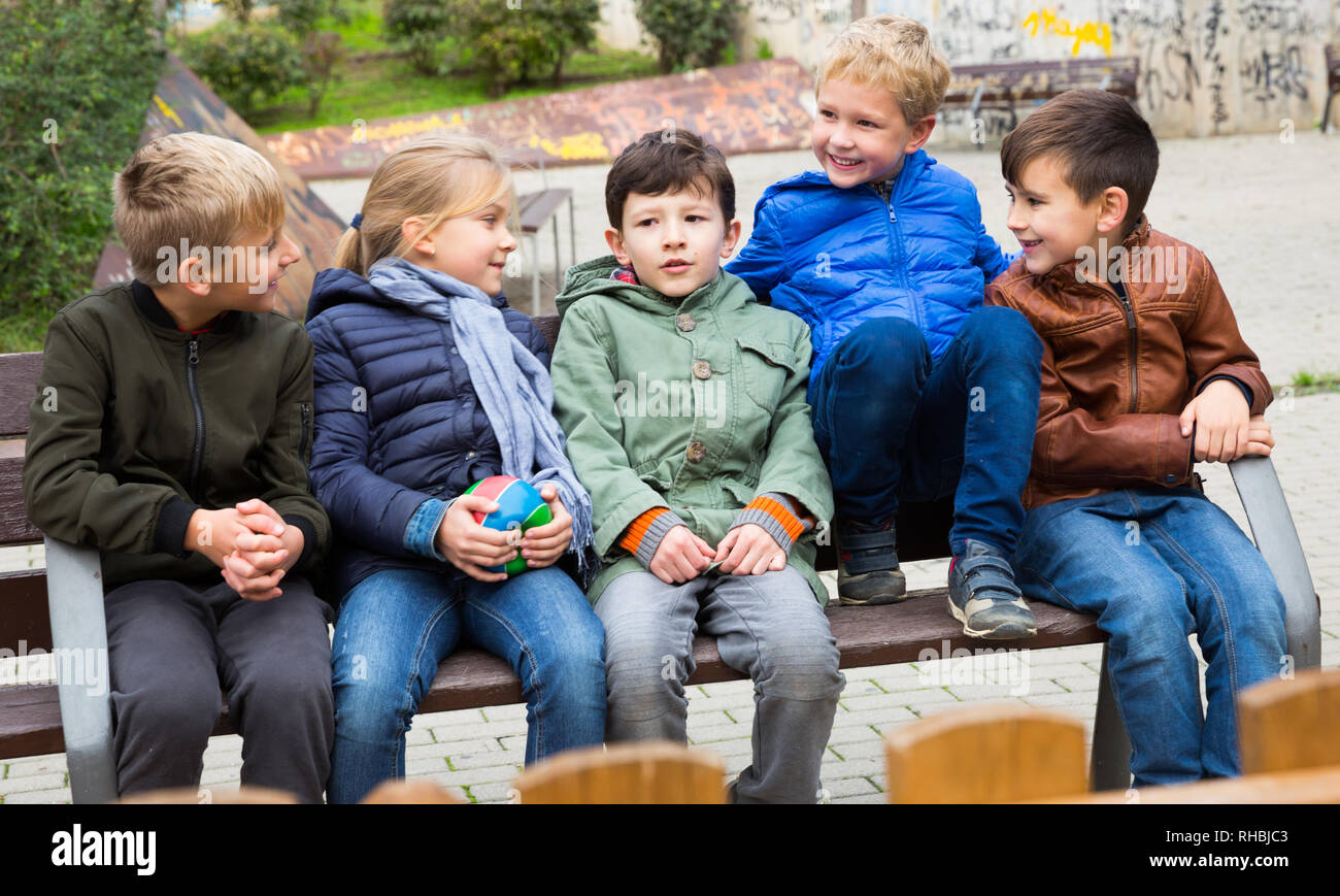 Group of cheerful children chatting while relaxing on bench on ...