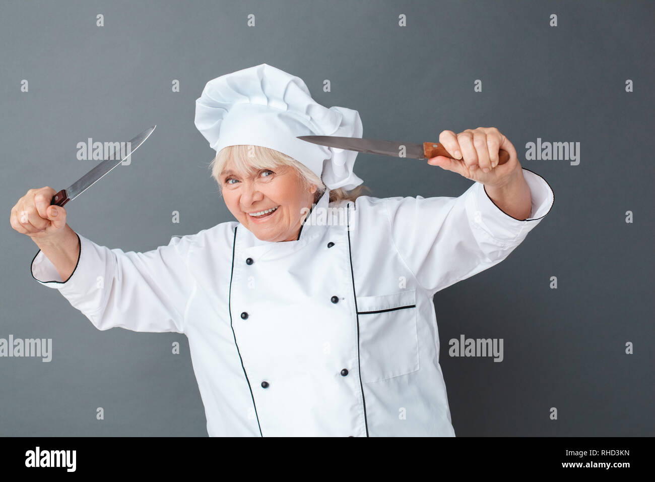Senior female chef studio standing isolated on gray wall holding knives ...