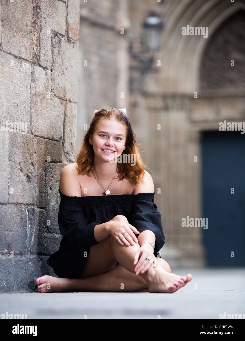 Positive young girl sitting barefoot near the stone wall at the street ...