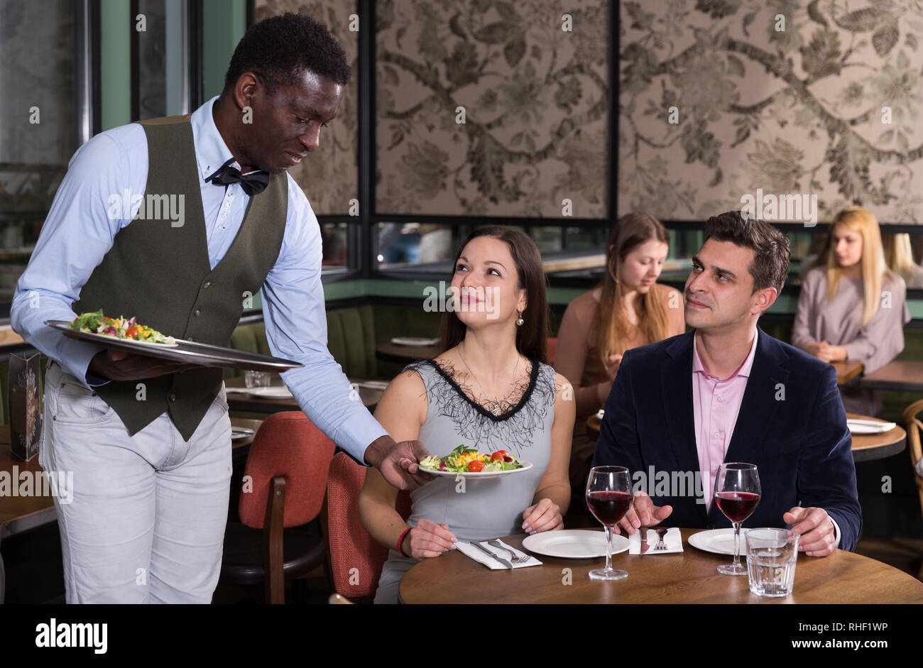 Polite African American waiter bringing ordered dishes to smiling ...