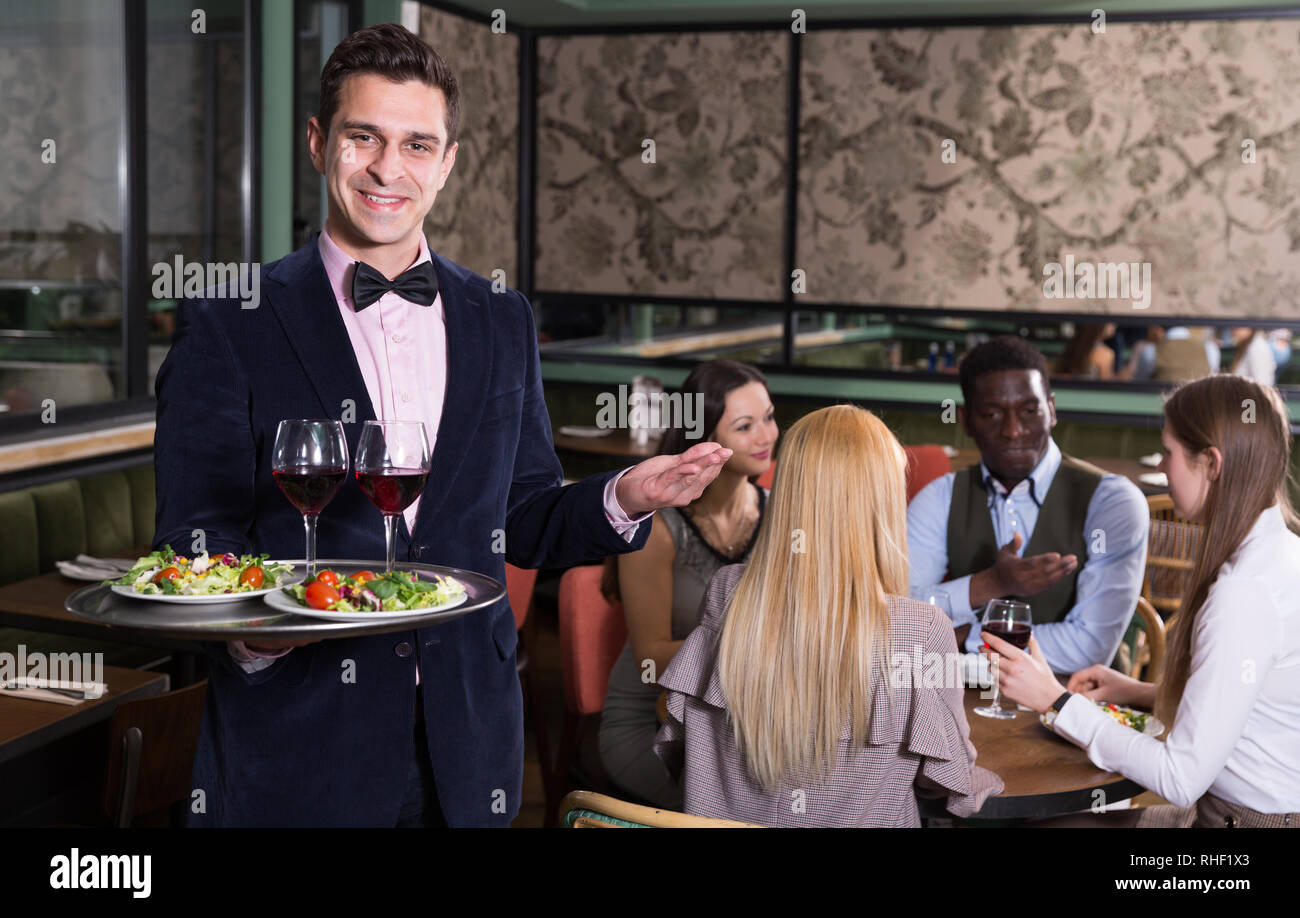 Polite waiter holding tray at restaurant with customers behind him ...