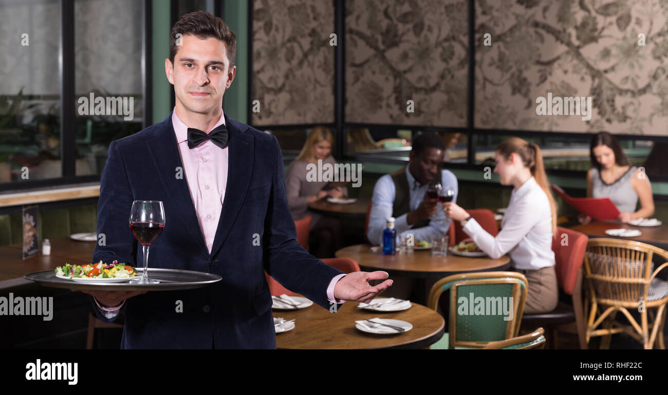 Polite waiter holding tray at restaurant with customers behind him ...
