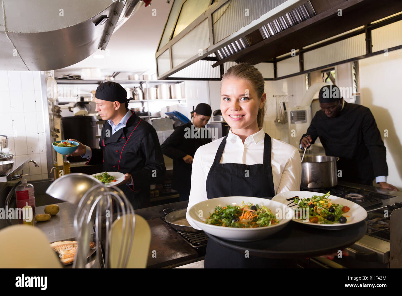 Portrait of attractive waitress with ordered dishes in kitchen of ...
