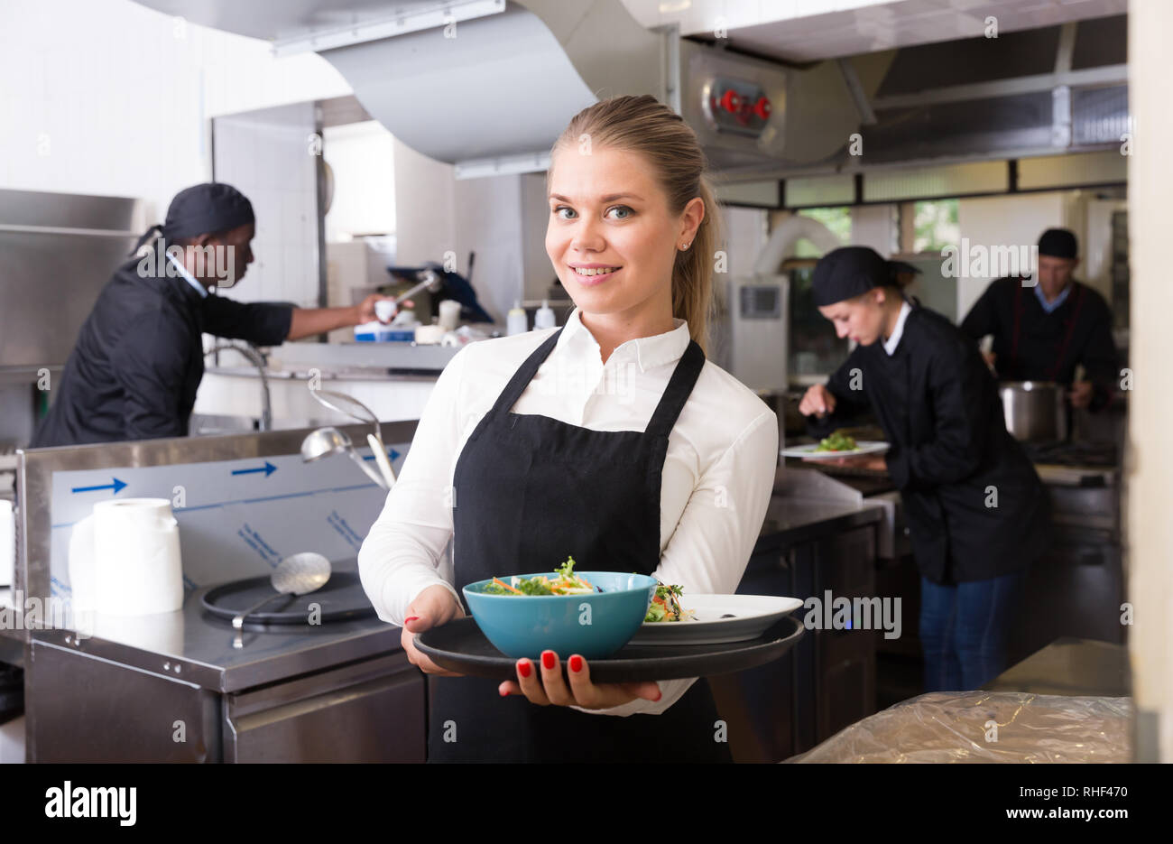 Portrait of attractive waitress with ordered dishes in kitchen of ...