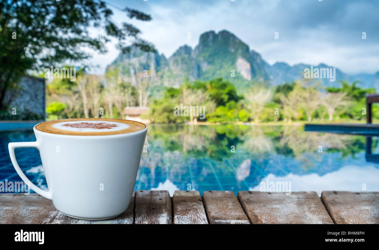 A white cup of coffee on the wooden table by the pool with mountain ...