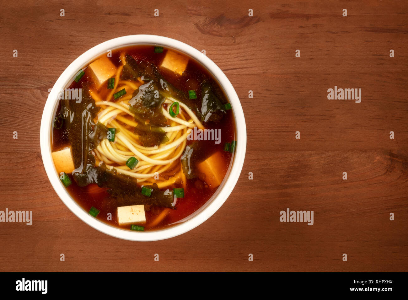 A bowl of miso shiru soup with tofu, scallions, noodles, and wakame seaweed, shot from the top ...