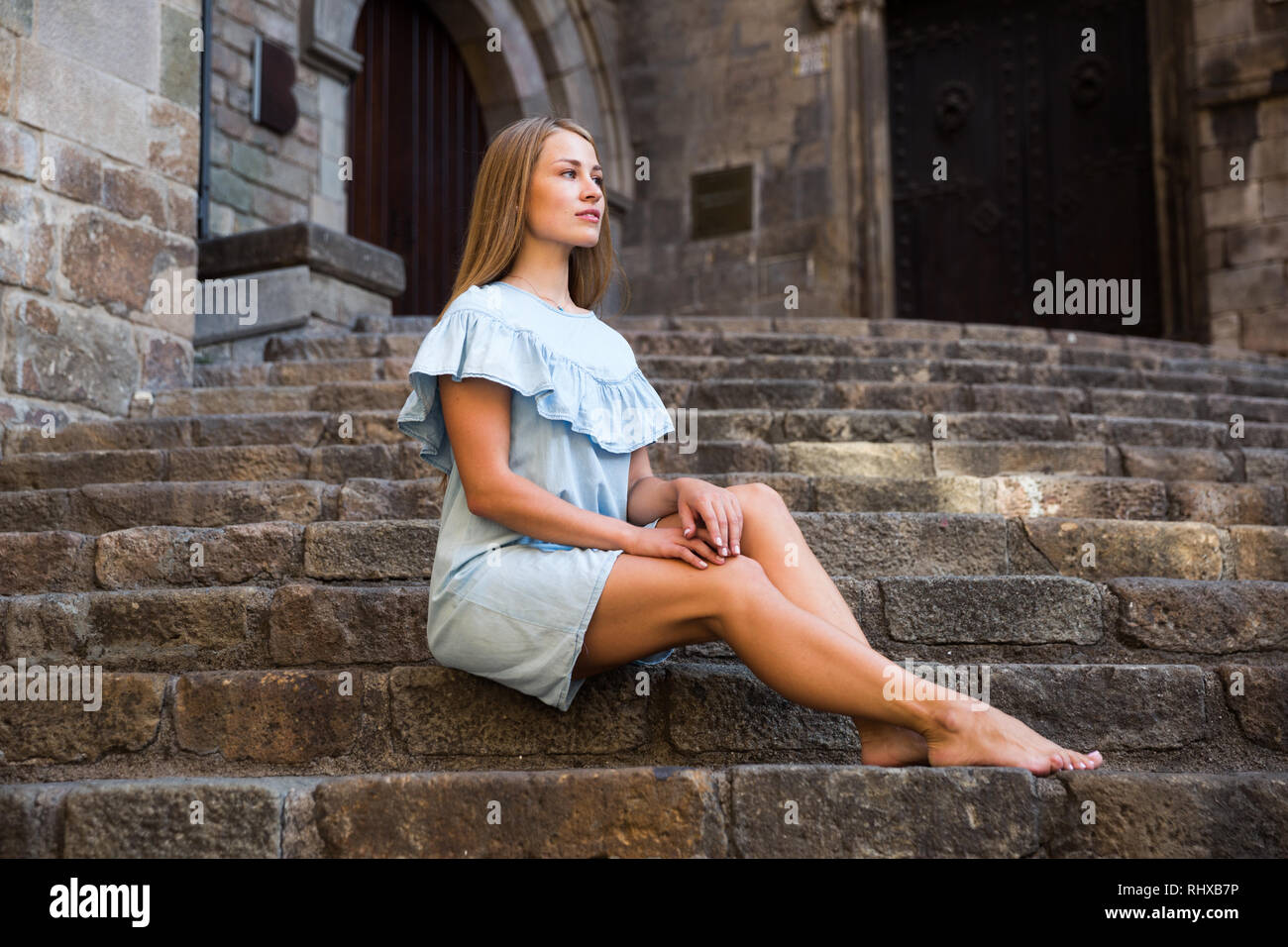Young smiling girl in lightweight blue dress sitting on stairs of temple in graceful pose in ...