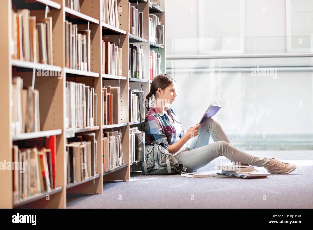 Student reading book while sitting against bookshelf at library Stock ...