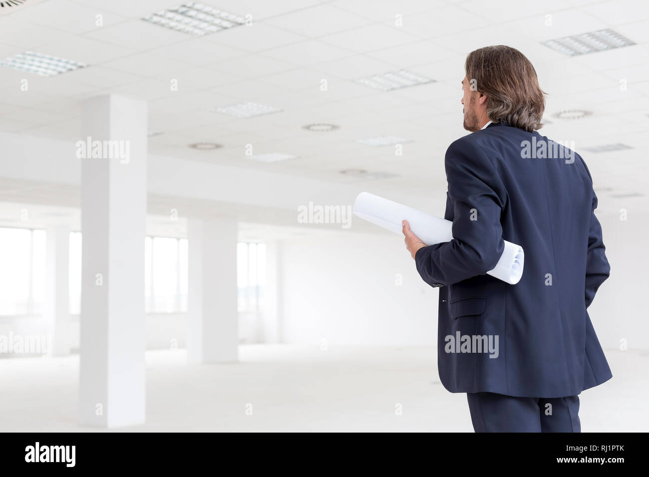 Rear view of businessman holding blueprint while standing in new empty ...