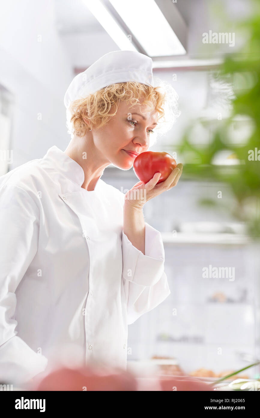Confident blond chef smelling fresh red tomato in kitchen at restaurant ...