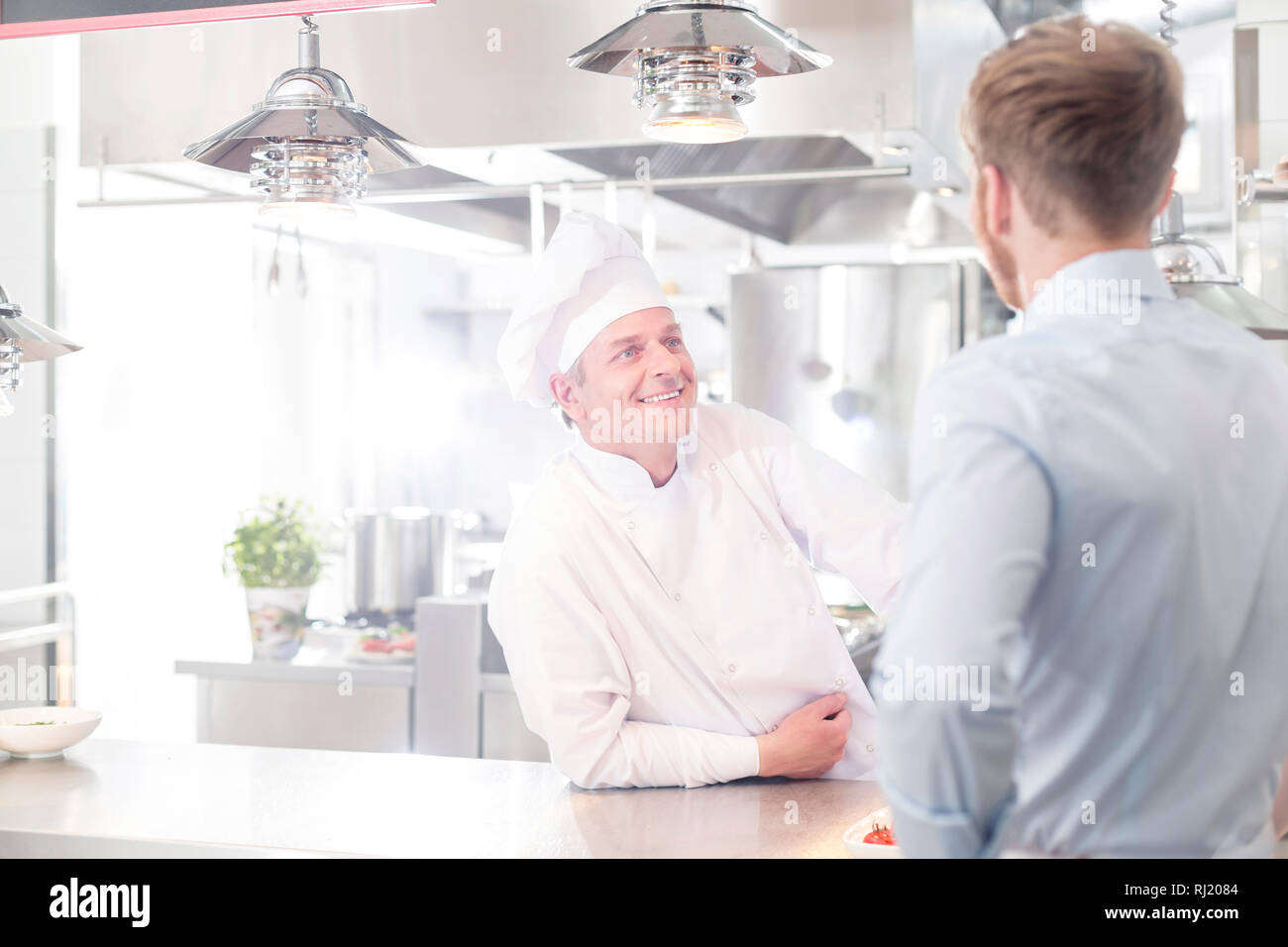 Smiling mature chef talking with young waiter at kitchen counter in ...