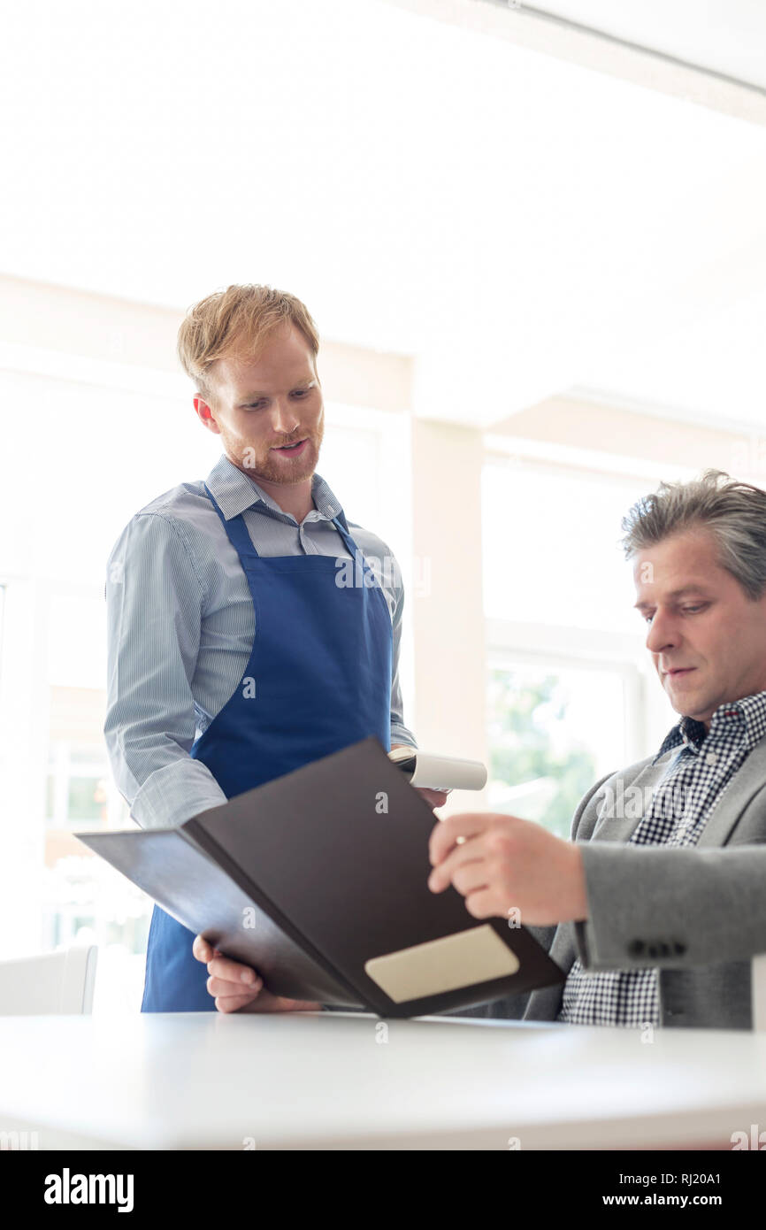 Young waiter standing by customer reading menu at table in restaurant ...