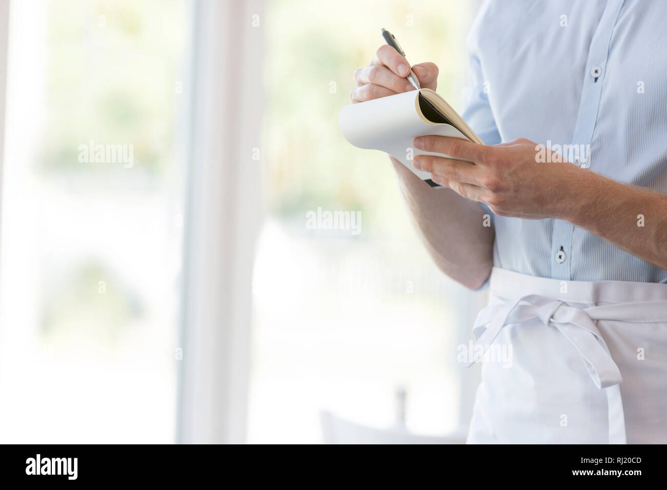 Midsection of young waiter writing on notepad while standing at ...