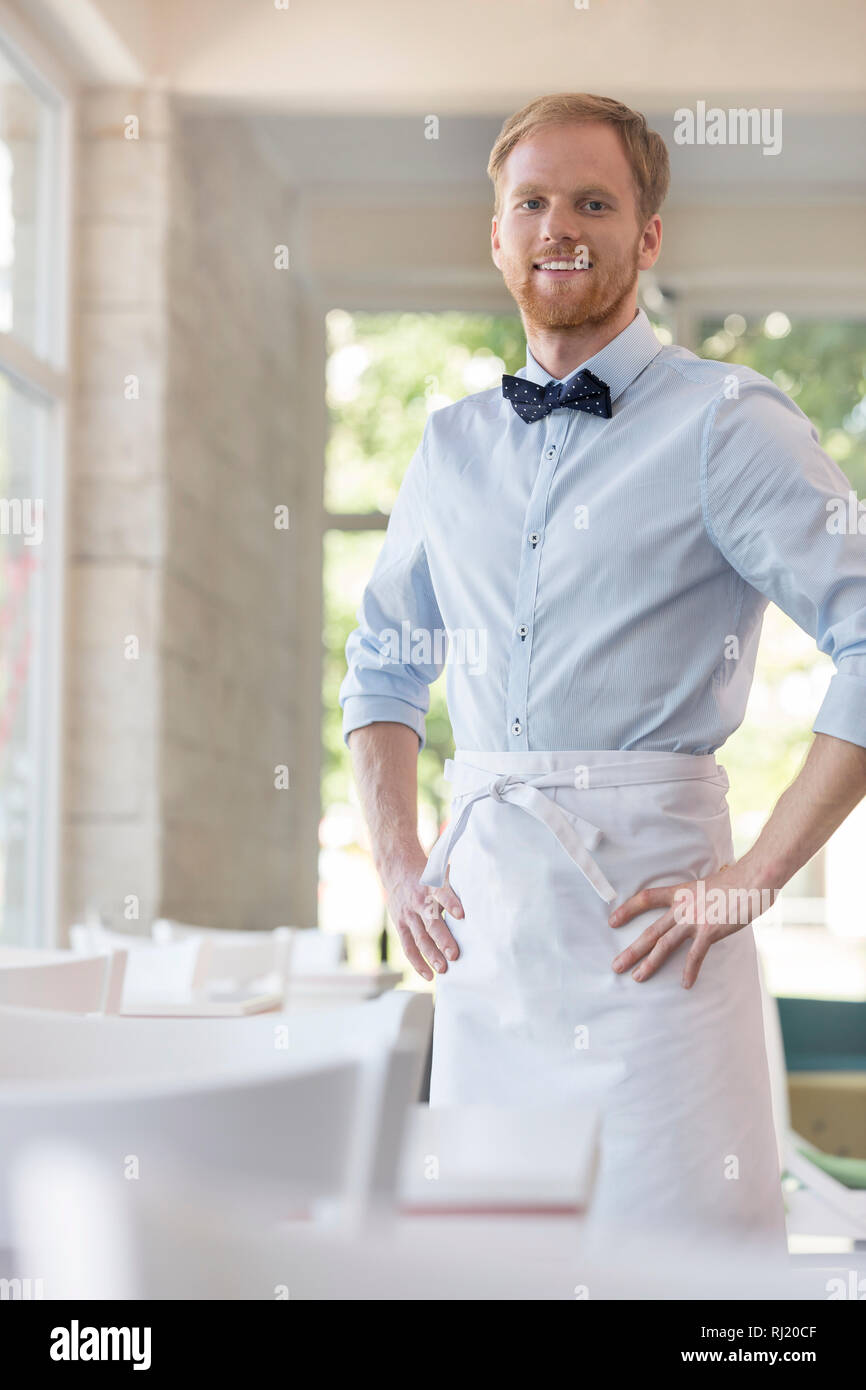 Portrait of smiling young waiter standing amidst furniture at ...