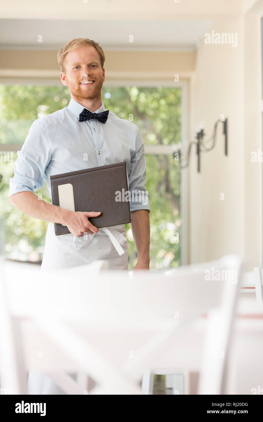 Portrait of confident young waiter holding menu while standing at ...