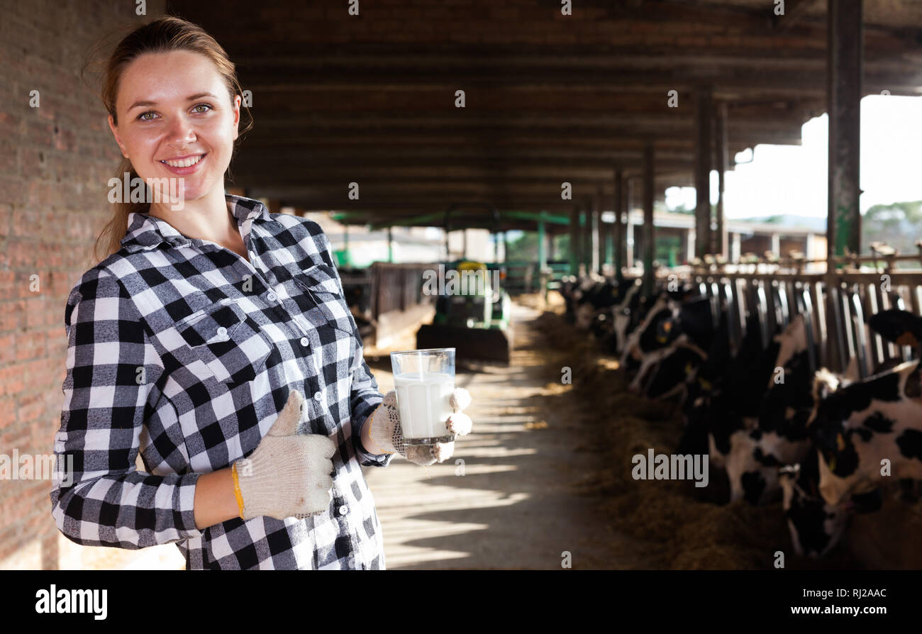 Young positive female dairy farm worker standing with glass of fresh ...