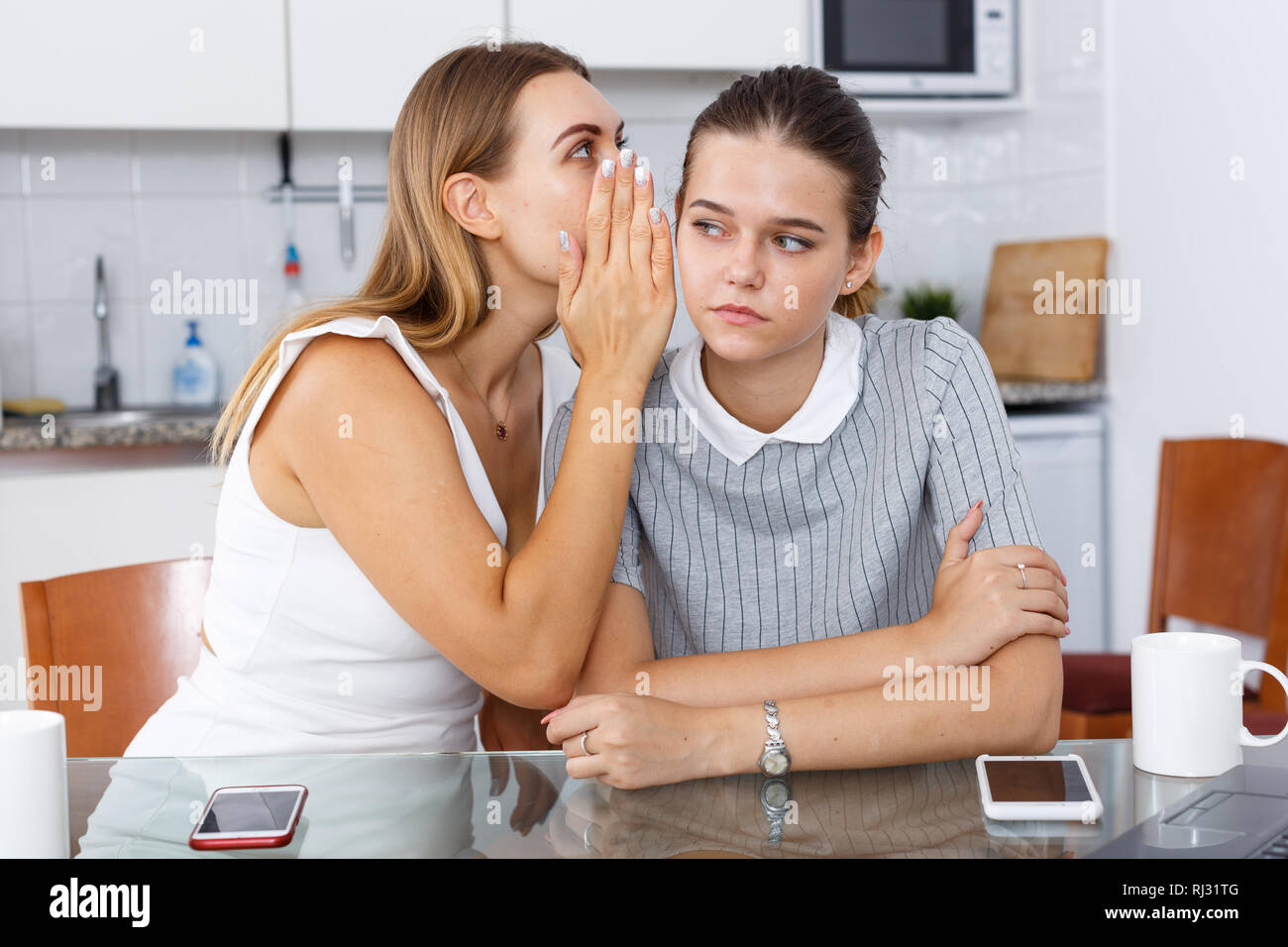 Two young girls friends whispering in kitchen interior Stock Photo - Alamy